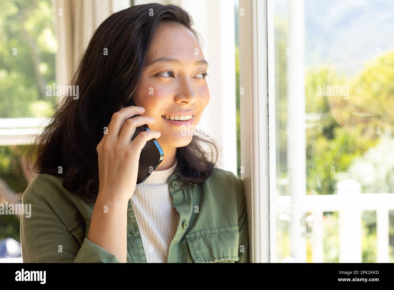 Happy asian woman standing next to window and talking on smartphone ...