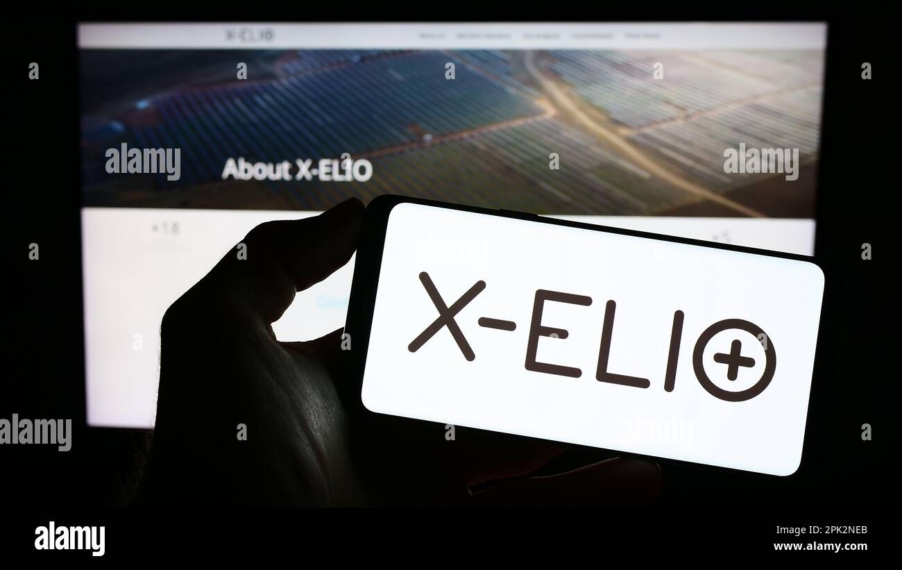 Person holding mobile phone with logo of Spanish renewables company X-ELIO Energy S.L. on screen ...