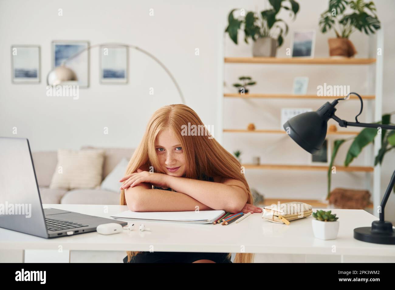 Leaning on the table. Female teenager with blonde hair is at home at ...