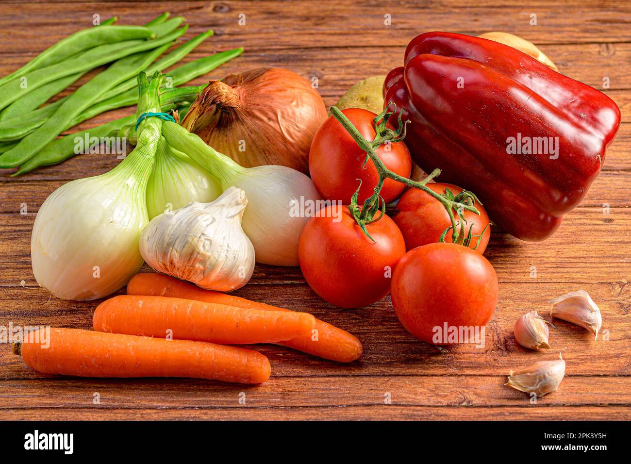 A rustic wooden table displaying a variety of fresh vegetables including tomatoes, beans, onions ...