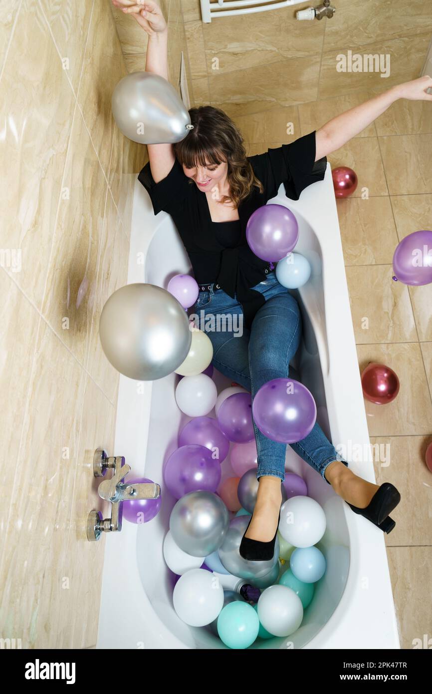 Young Woman Sitting in a Bathtub Full of Balloons in Bathroom Throwing ...