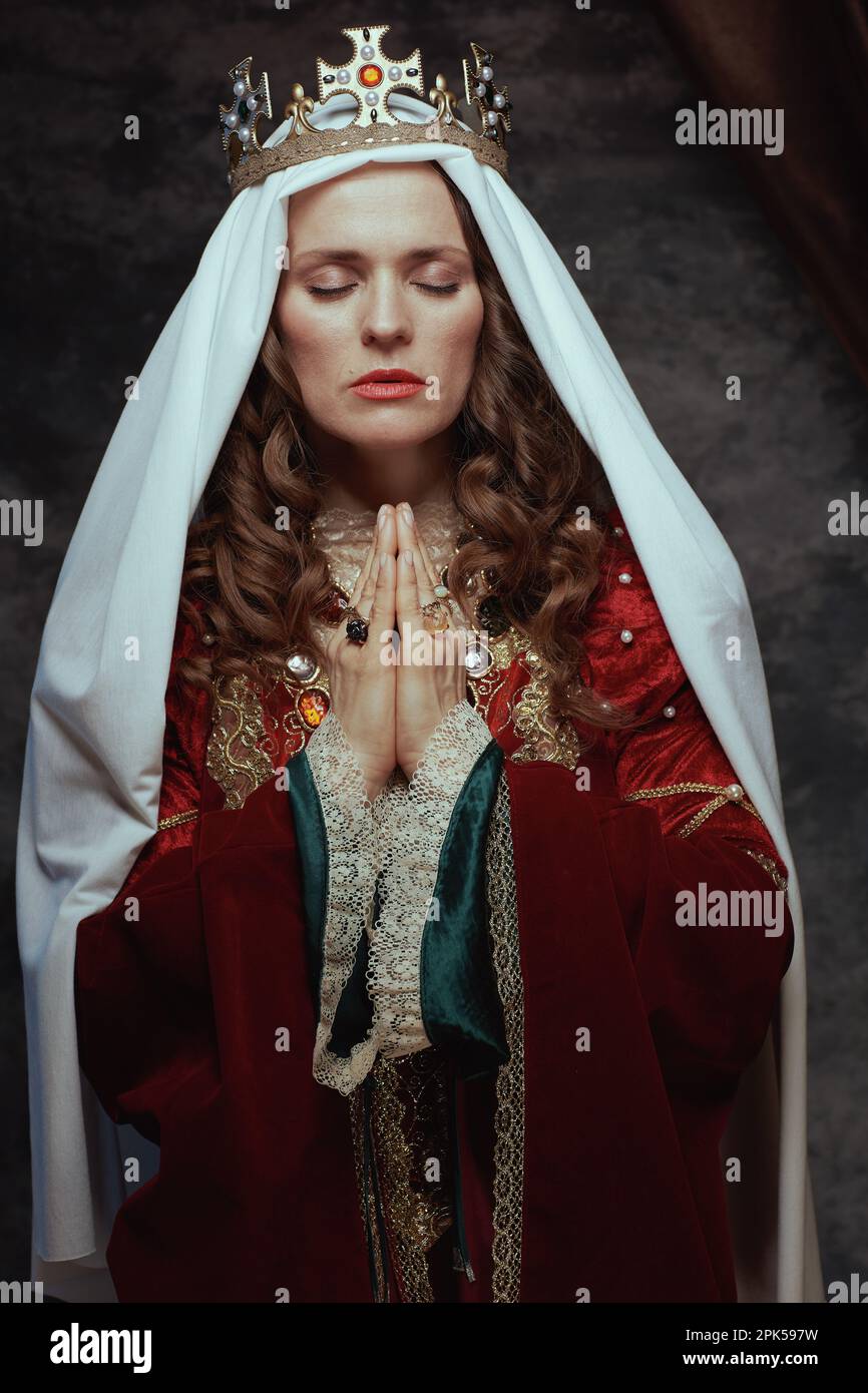 medieval queen in red dress with veil and crown praying on dark gray ...