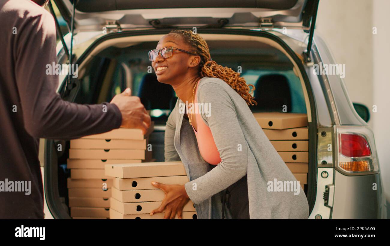 Female courier delivering pizza boxes stack from car trunk, taking huge pizzeria order in piles ...
