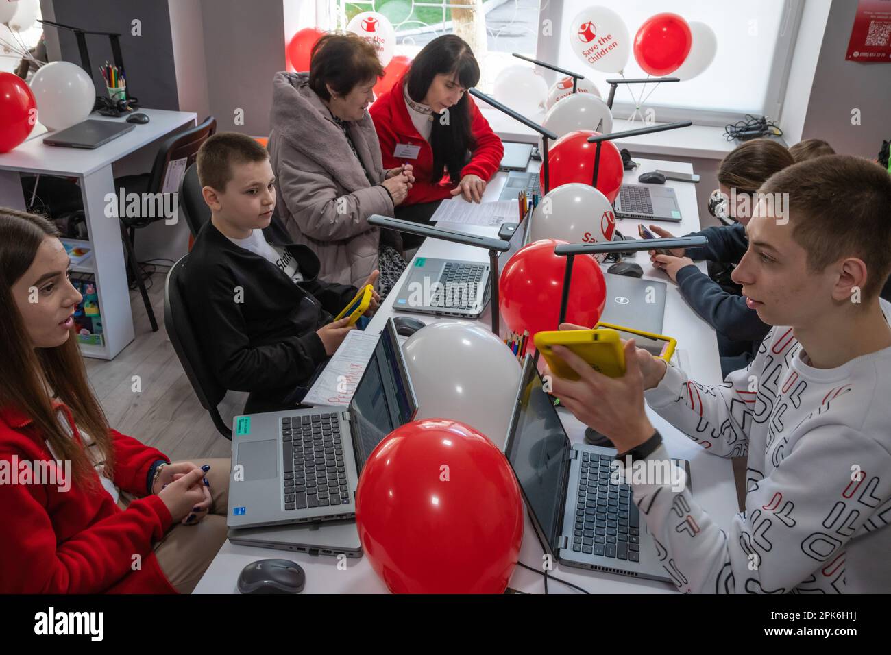 Children sit at a table with laptops and tablets in a classroom ...