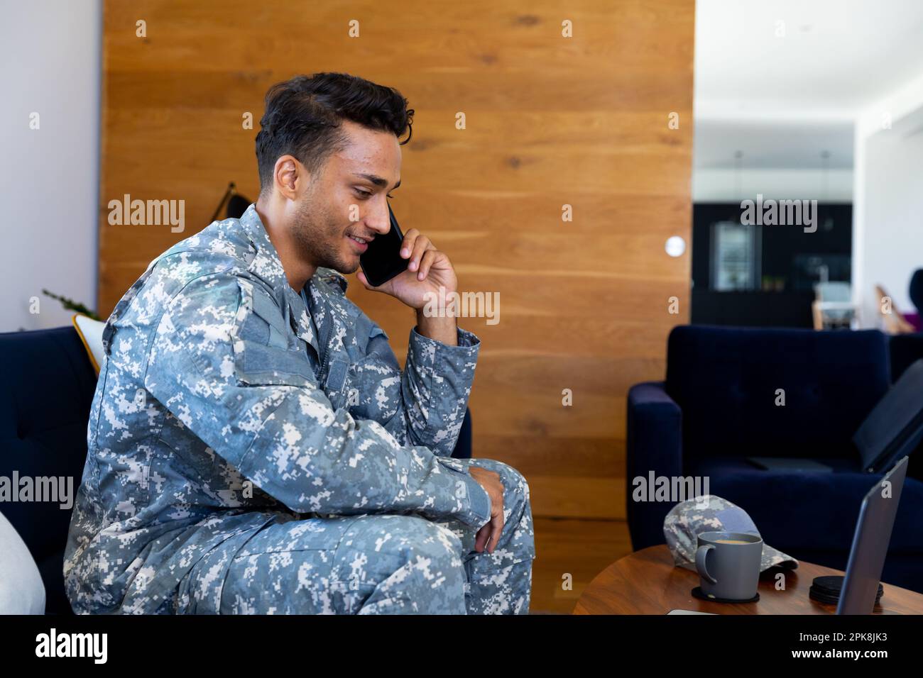 Happy biracial male soldier wearing uniform, sitting on sofa talking on ...