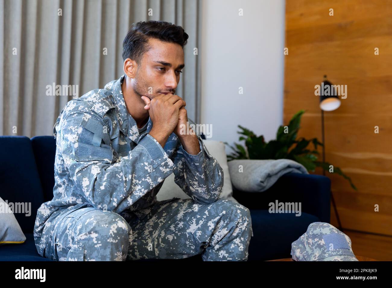 Thoughtful biracial male soldier wearing uniform, sitting on sofa in ...