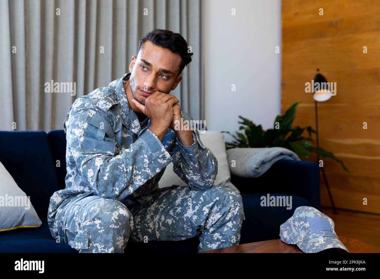 Thoughtful biracial male soldier wearing uniform, sitting on sofa in ...
