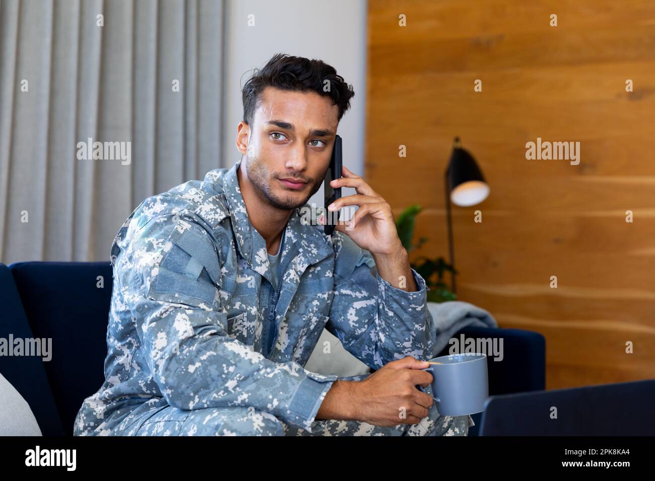 Thoughtful biracial male soldier in uniform, sitting on sofa talking on ...