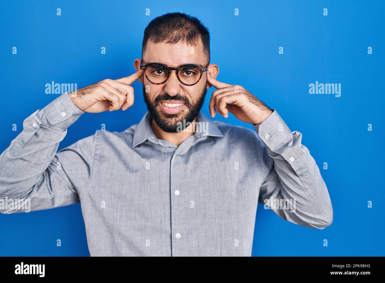 Middle east man with beard standing over blue background covering ears ...
