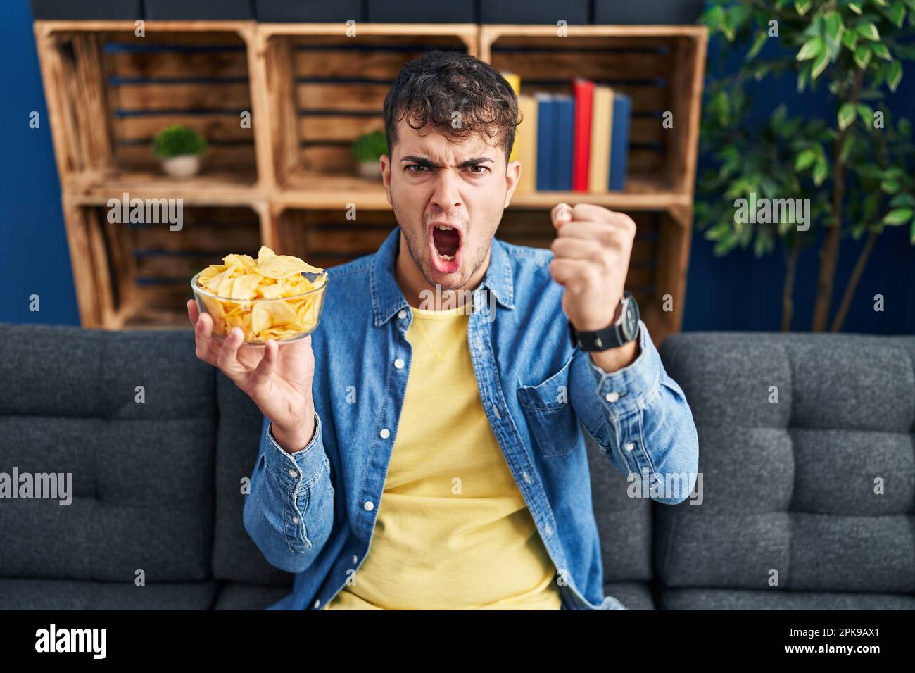 Young hispanic man holding potato chips annoyed and frustrated shouting ...