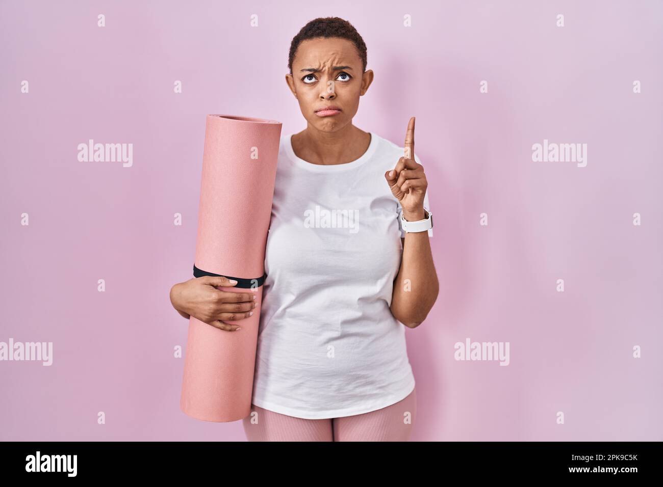 Beautiful african american woman holding yoga mat pointing up looking ...