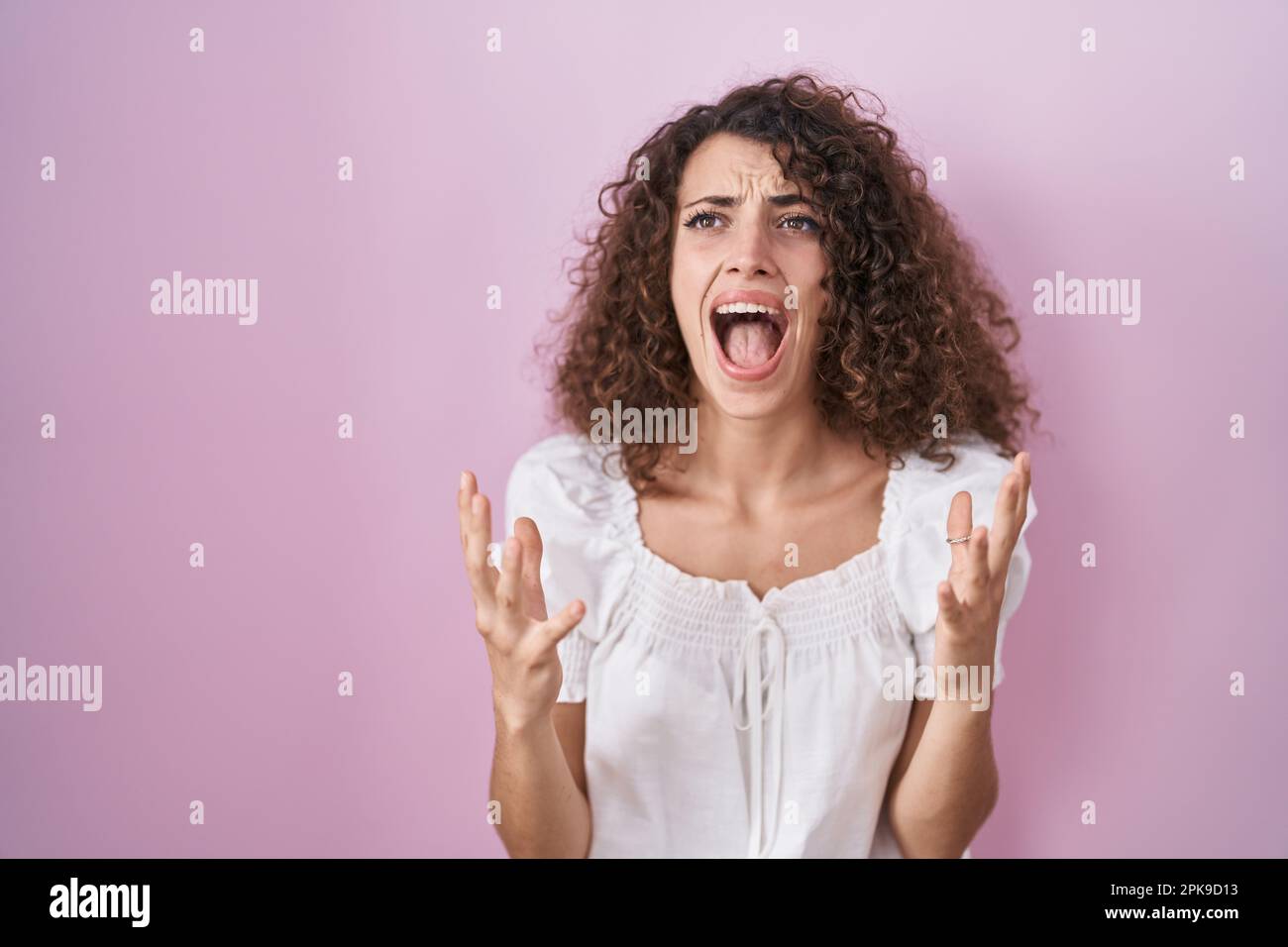 Hispanic woman with curly hair standing over pink background crazy and ...