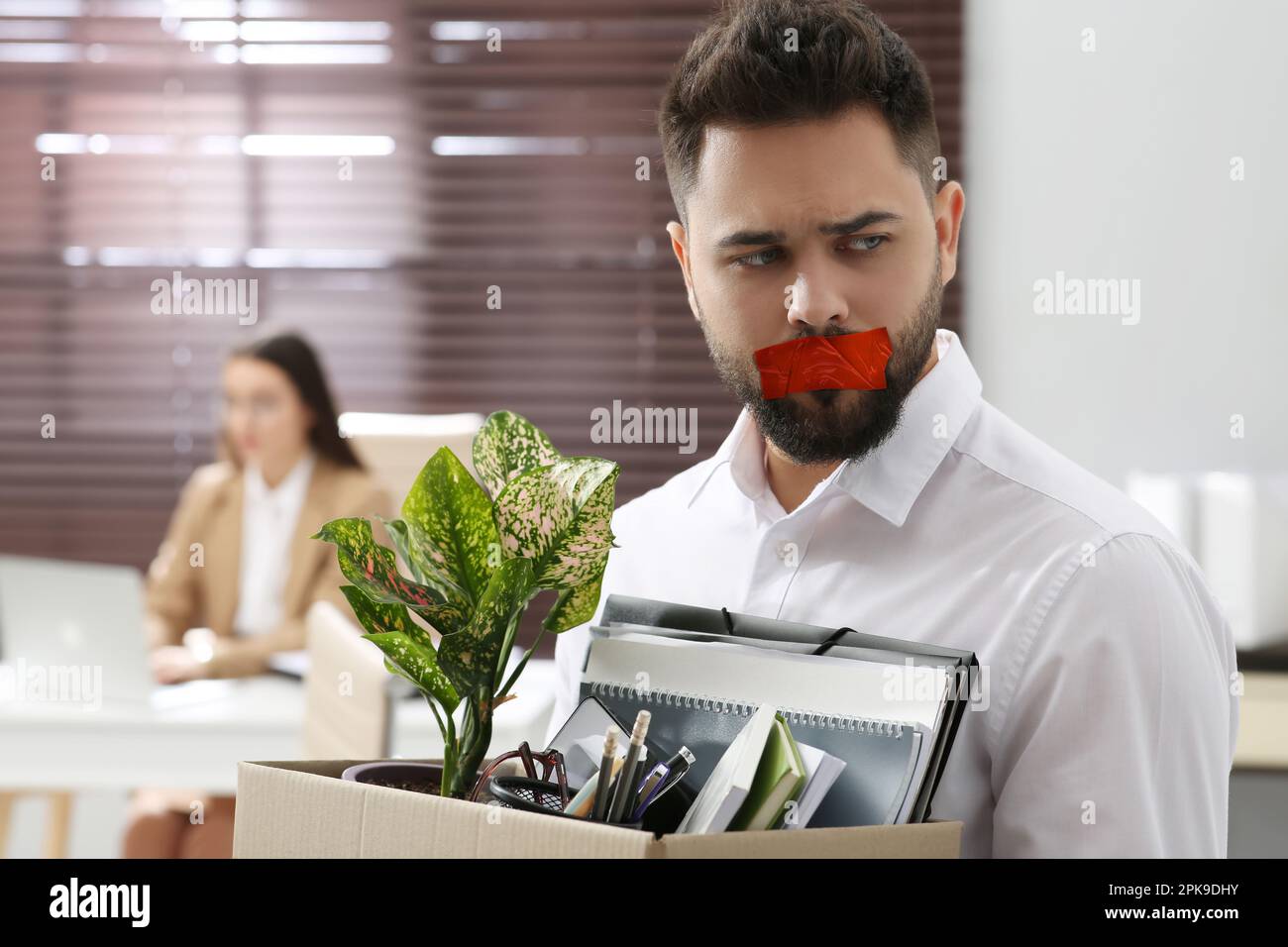 Upset dismissed man with taped mouth carrying box with personal stuff ...