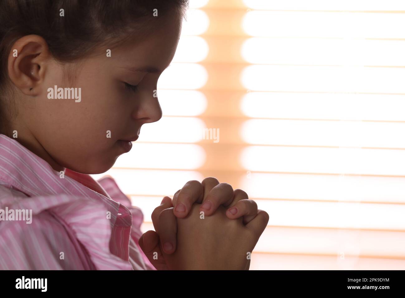 Cute little girl with hands clasped together praying near window ...