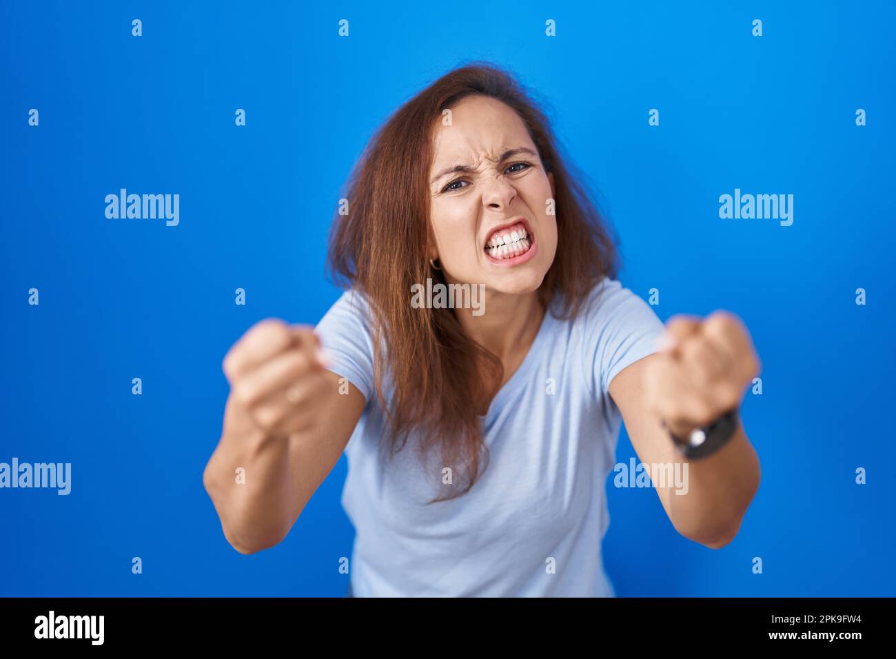 Brunette woman standing over blue background angry and mad raising ...