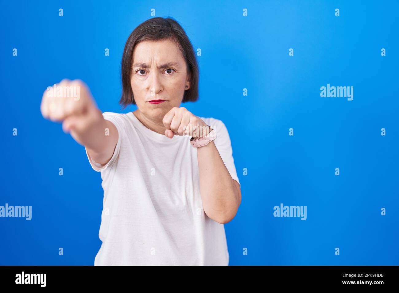 Middle age hispanic woman standing over blue background punching fist ...