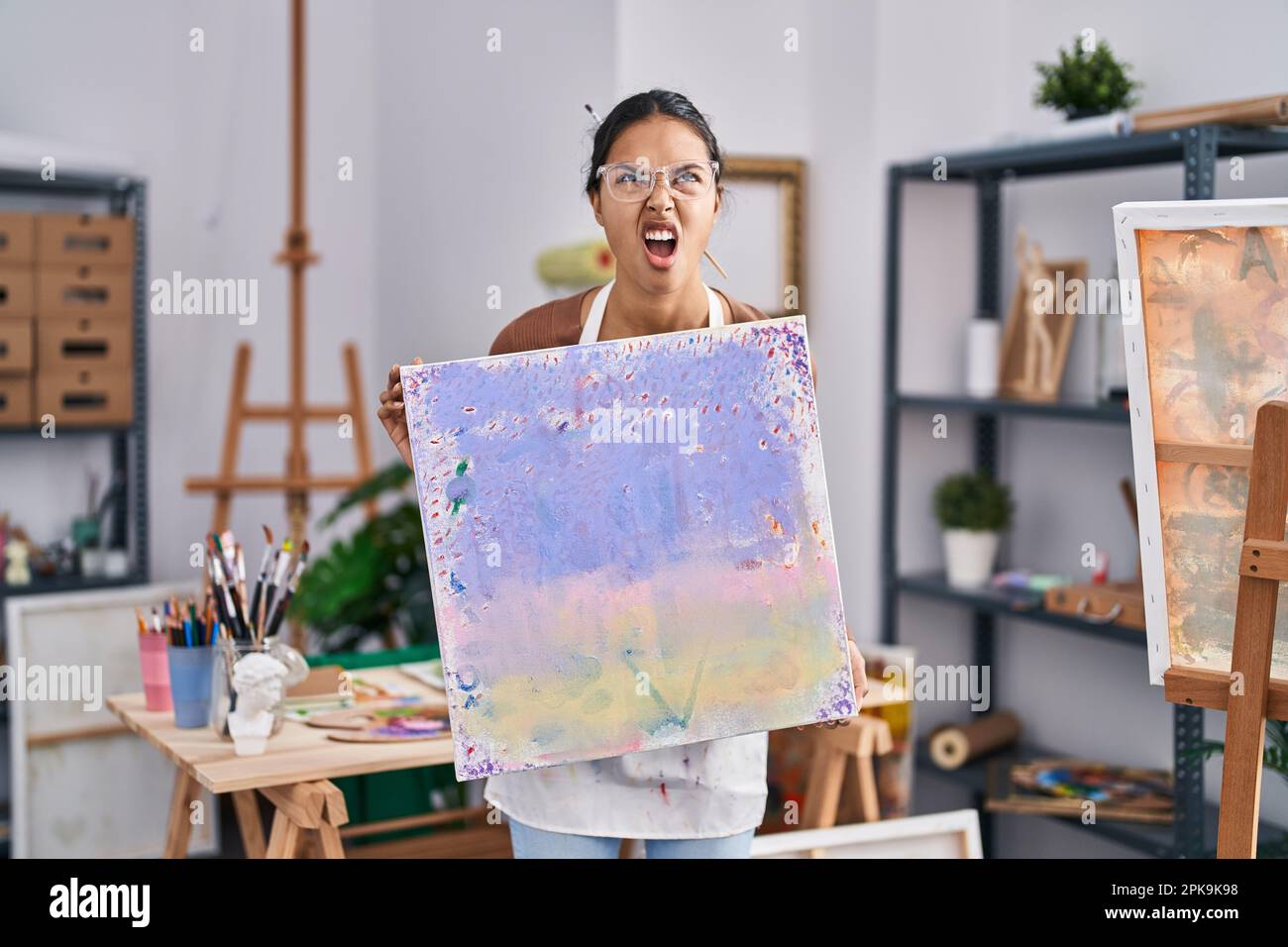 Young brazilian woman holding painter canvas at art studio angry and ...