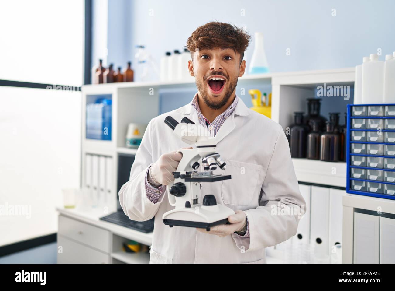 Arab man with beard working at scientist laboratory holding microscope celebrating crazy and ...