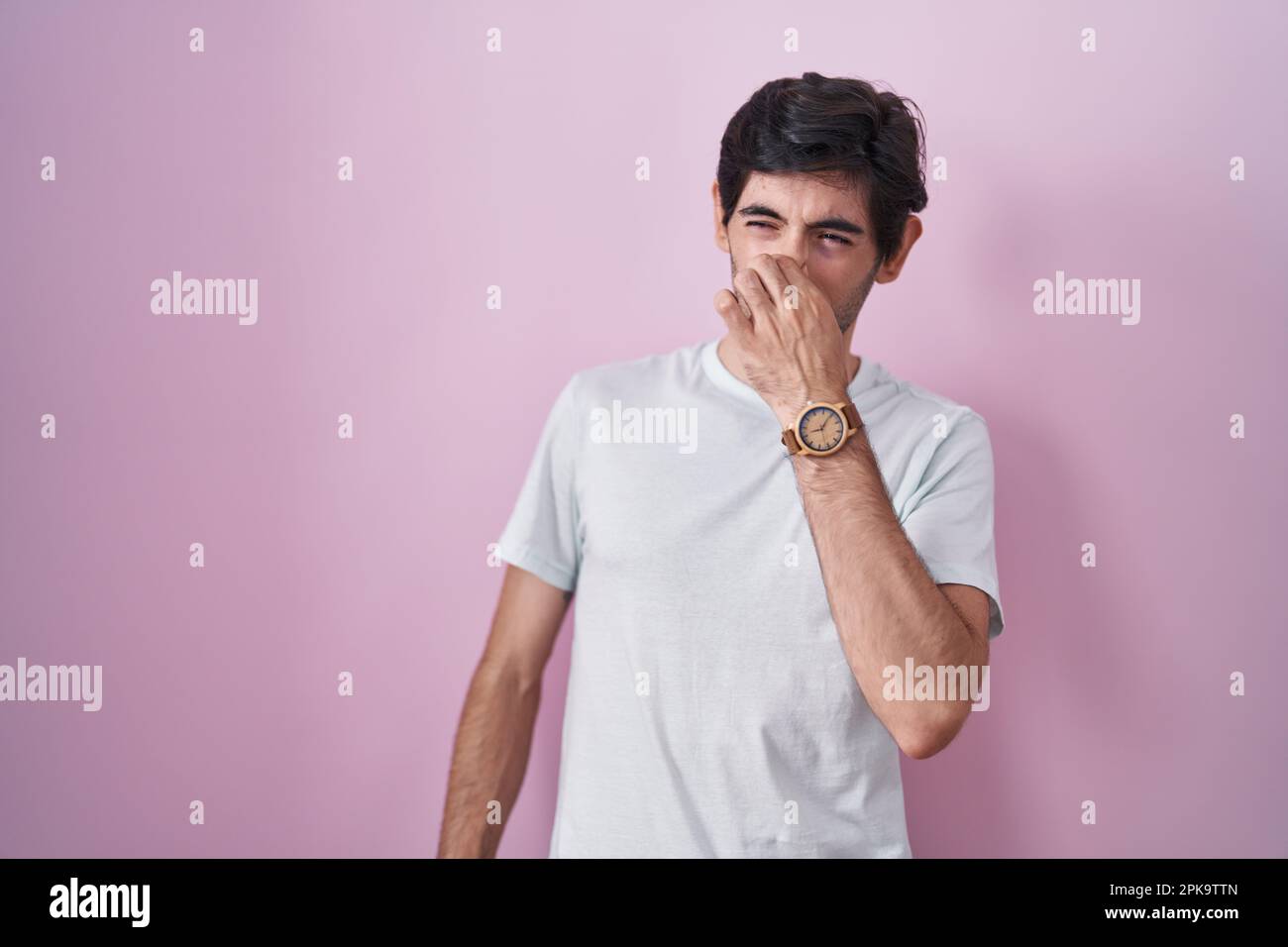 Young hispanic man standing over pink background smelling something ...
