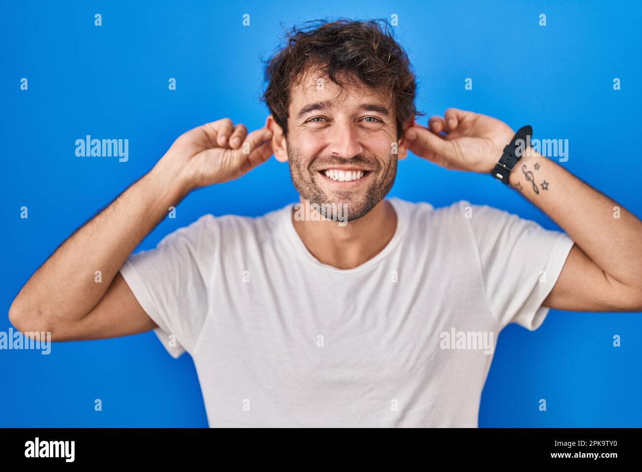 Hispanic young man standing over blue background smiling pulling ears ...