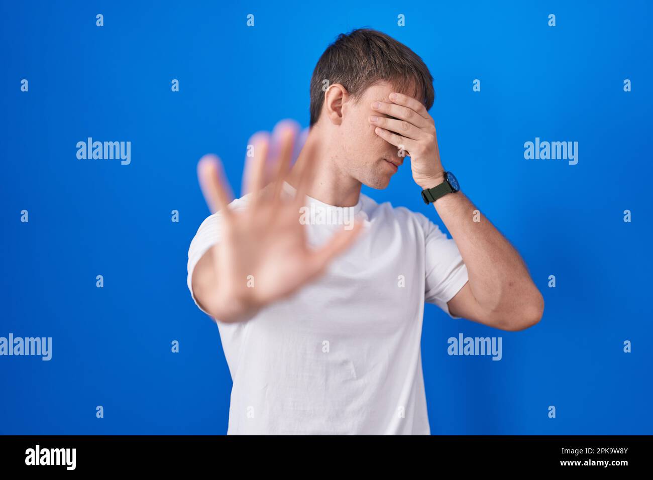 Caucasian blond man standing over blue background covering eyes with ...