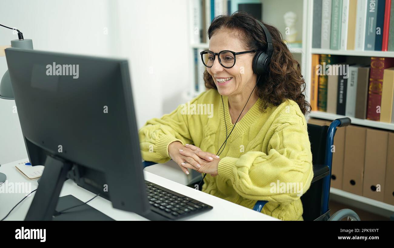 Middle age hispanic woman teacher using computer sitting on wheelchair at library university ...