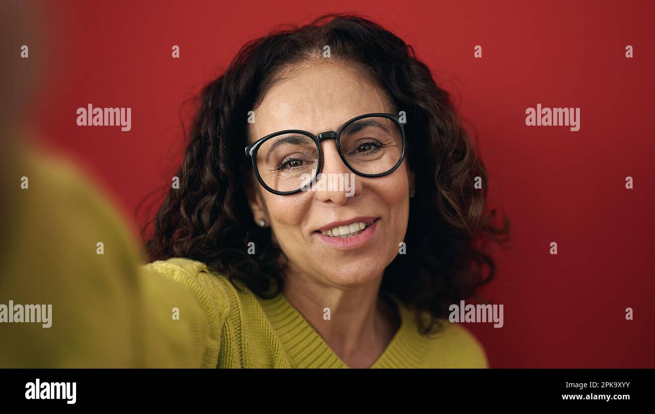 Middle age hispanic woman smiling confident make selfie by camera over isolated red background ...