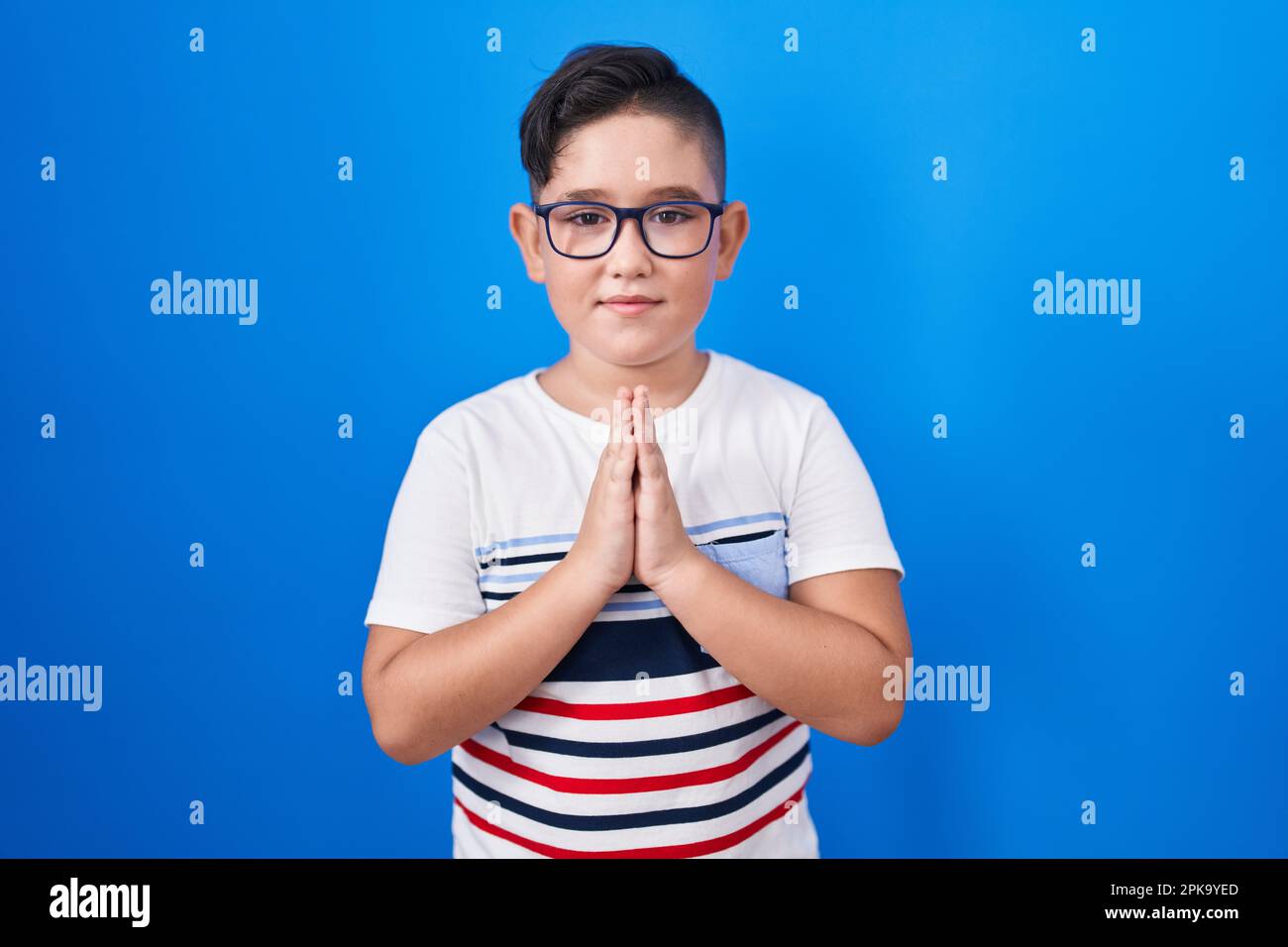Young hispanic kid standing over blue background praying with hands ...