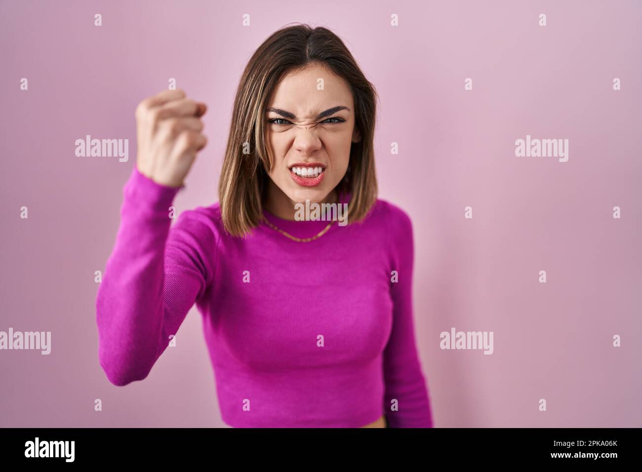 Hispanic woman standing over pink background angry and mad raising fist ...