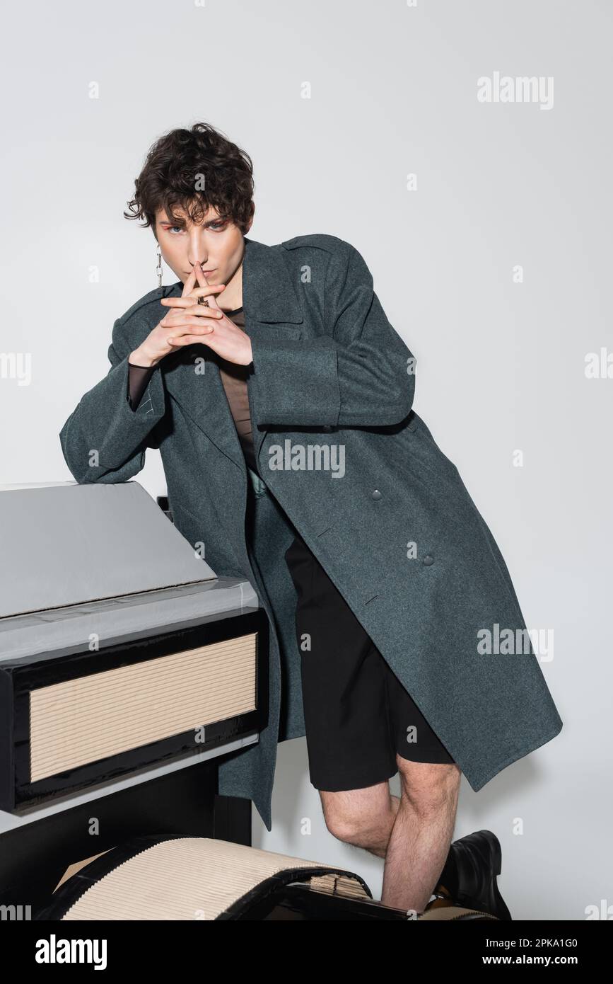 thoughtful pangender person in coat posing with clenched hands near ...