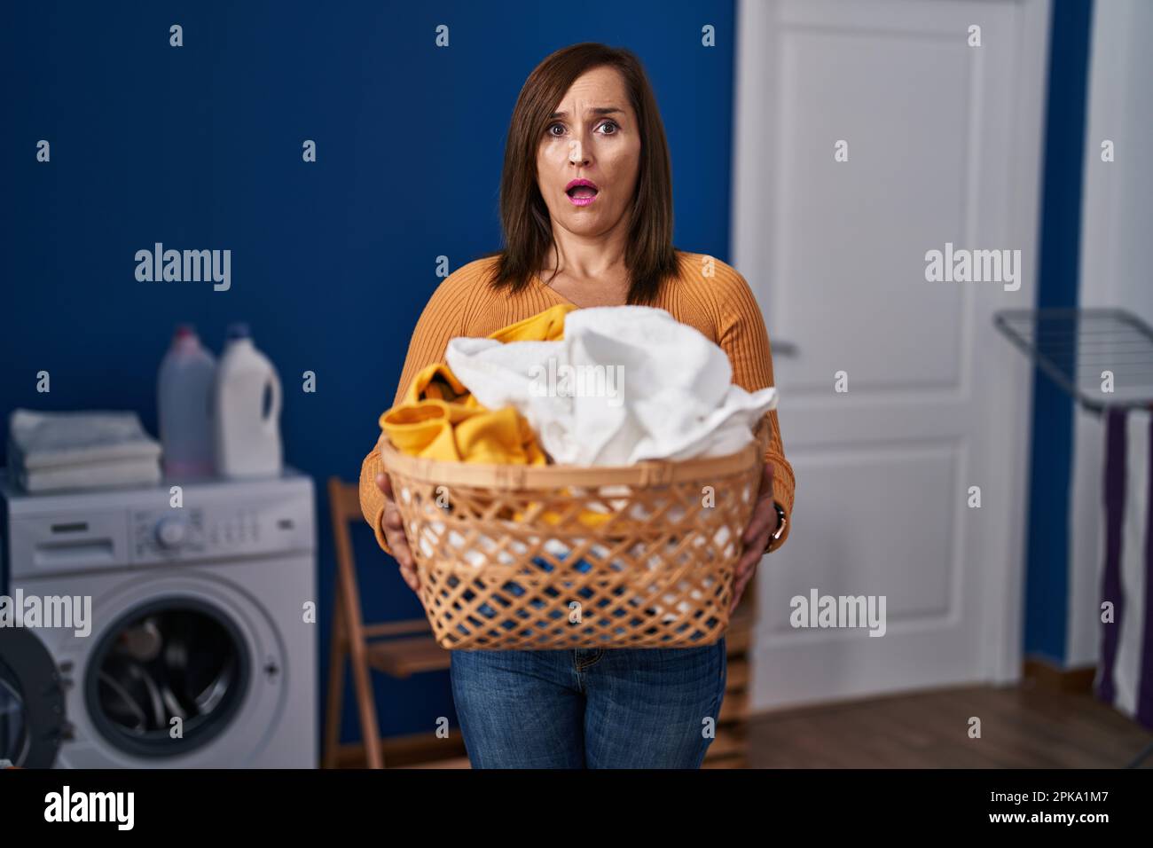 Middle age woman holding laundry basket at laundry room in shock face ...