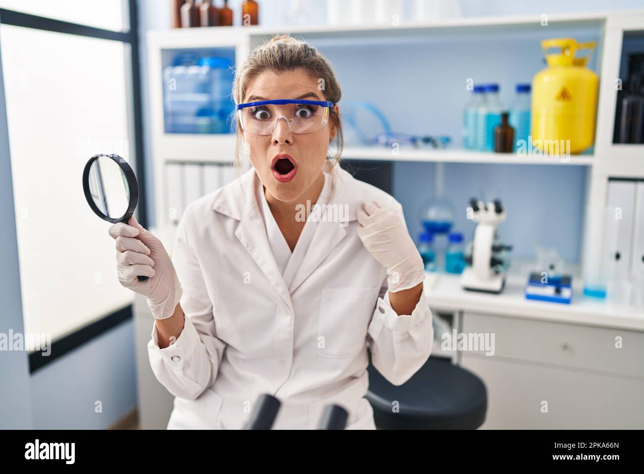 Young woman working at scientist laboratory holding magnifying glass ...