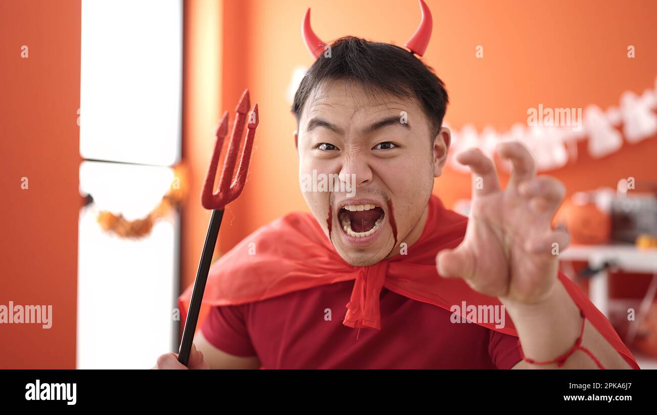 Young chinese man wearing devil costume doing scare gesture at home ...