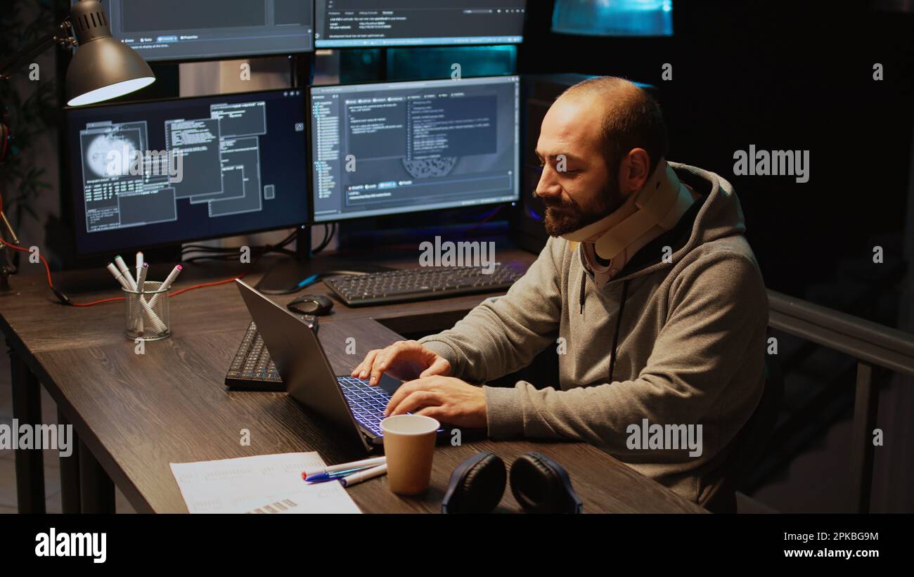 Young IT programmer wearing cervical collar at office, typing server code on multiple screens ...
