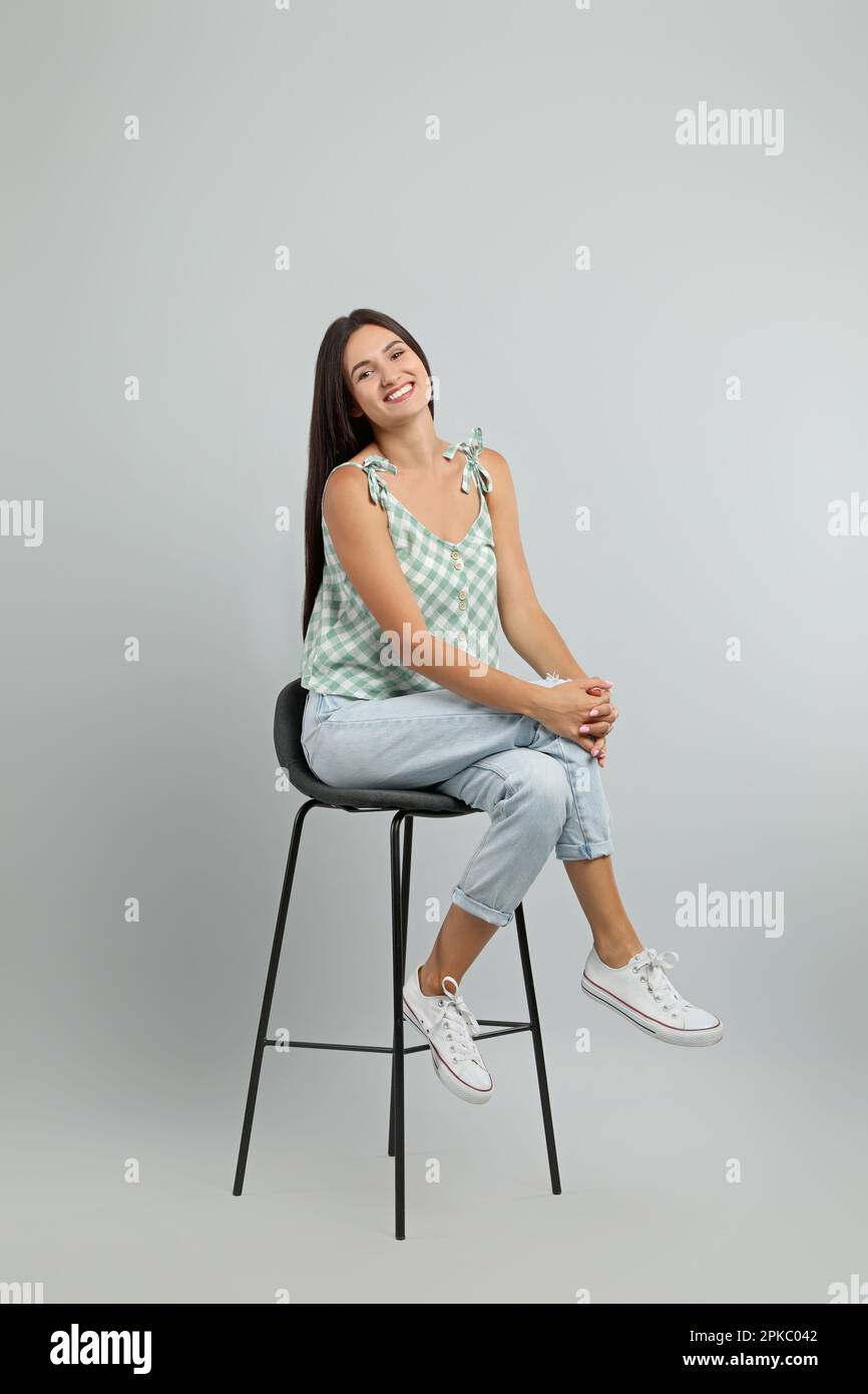 Beautiful young woman sitting on stool against light grey background ...