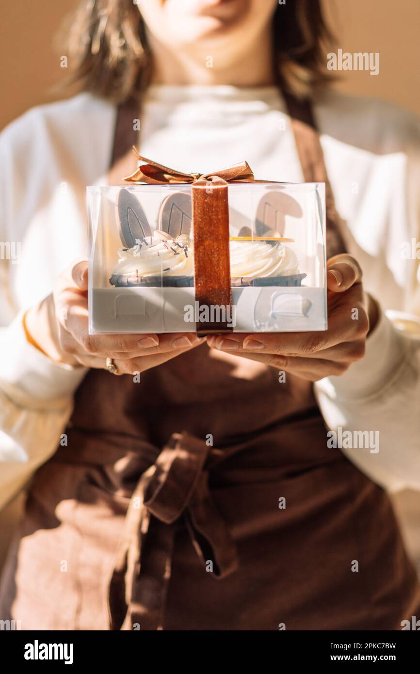 Lady pastry chef in brown apron holding box of cupcakes with bunny ears ...
