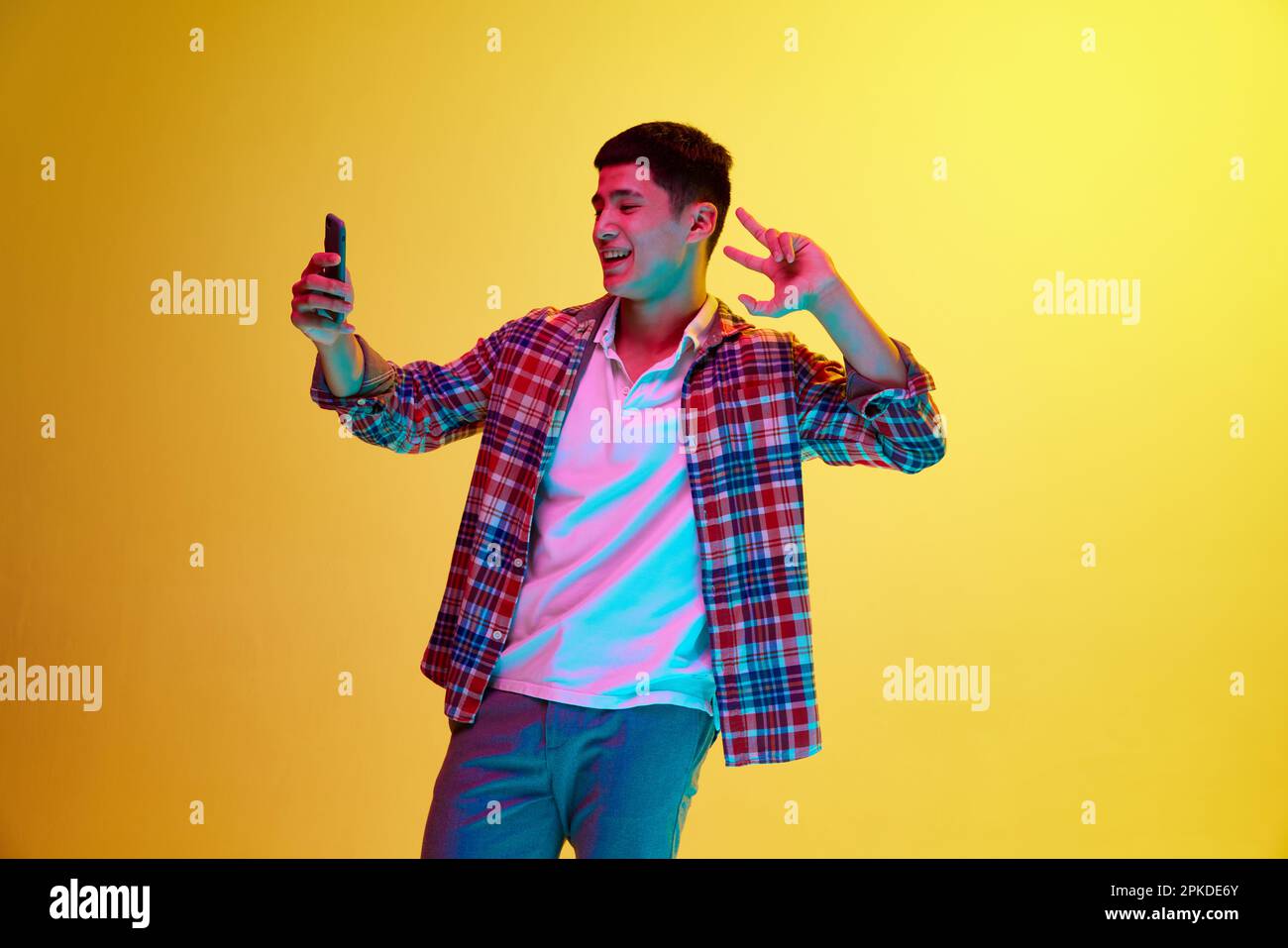 Positive, smiling, young guy taking selfie with mobile phone, posing against yellow background ...