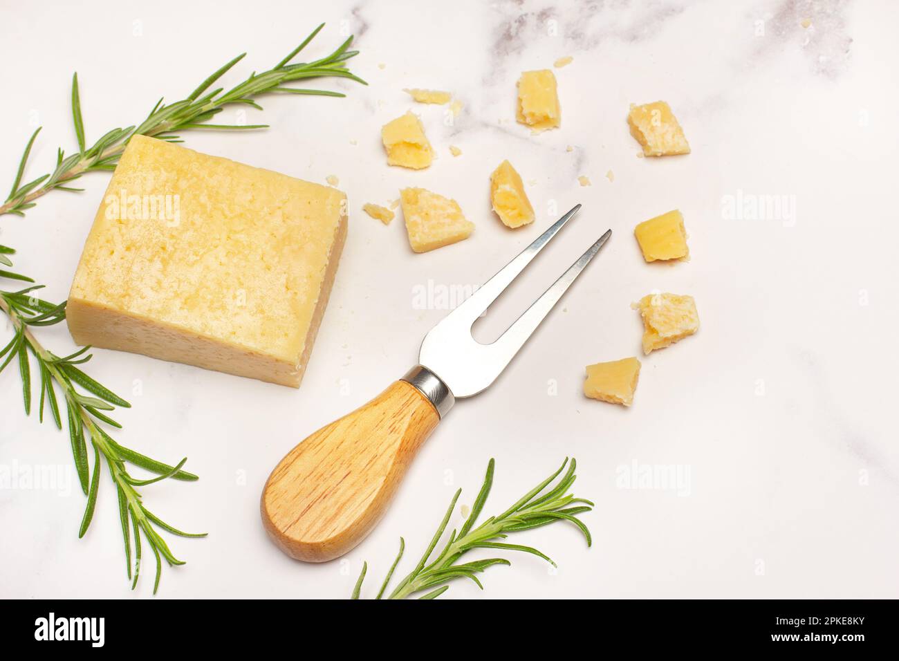 A piece of parmesan cheese, a fork and rosemary twigs on a white and ...
