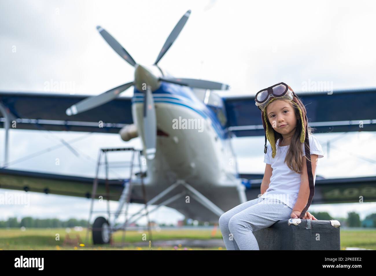 A little girl in a pilot's costume sits on a suitcase against the ...