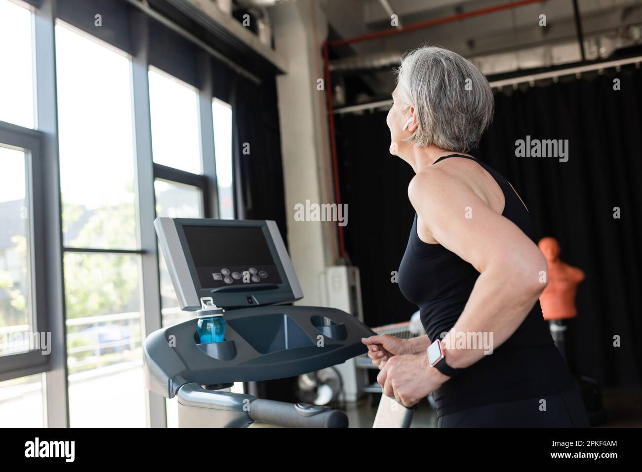 retired woman with grey hair running on treadmill next to sports bottle ...
