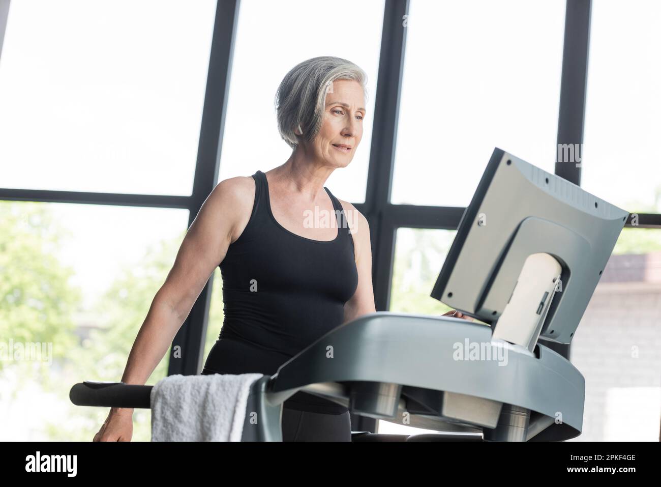 retired woman with grey hair looking at monitor of treadmill while ...