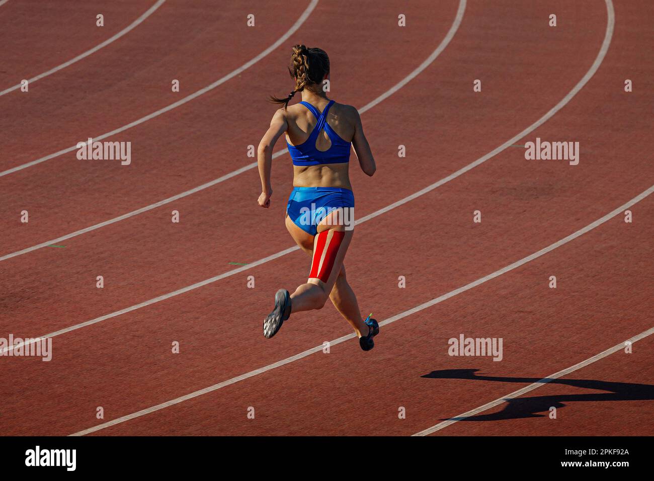 female runner running on red track stadium, kinesiotaping on back thigh ...