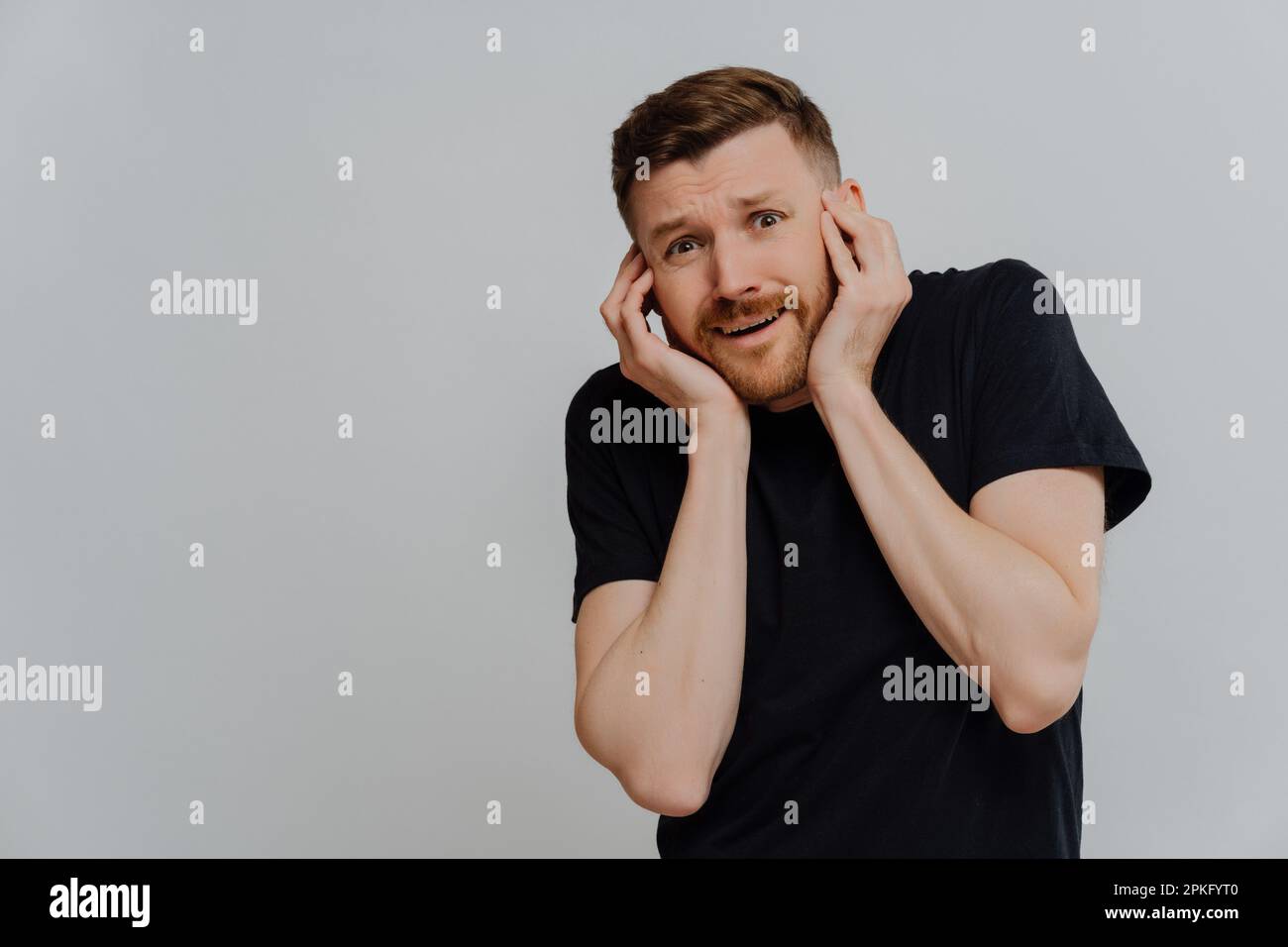 Scared young bearded man in black tshirt holding hands near face and ...