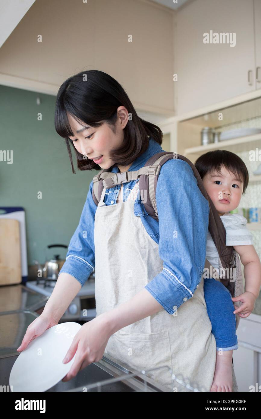 Mother washing dishes in kitchen with child on back Stock Photo - Alamy