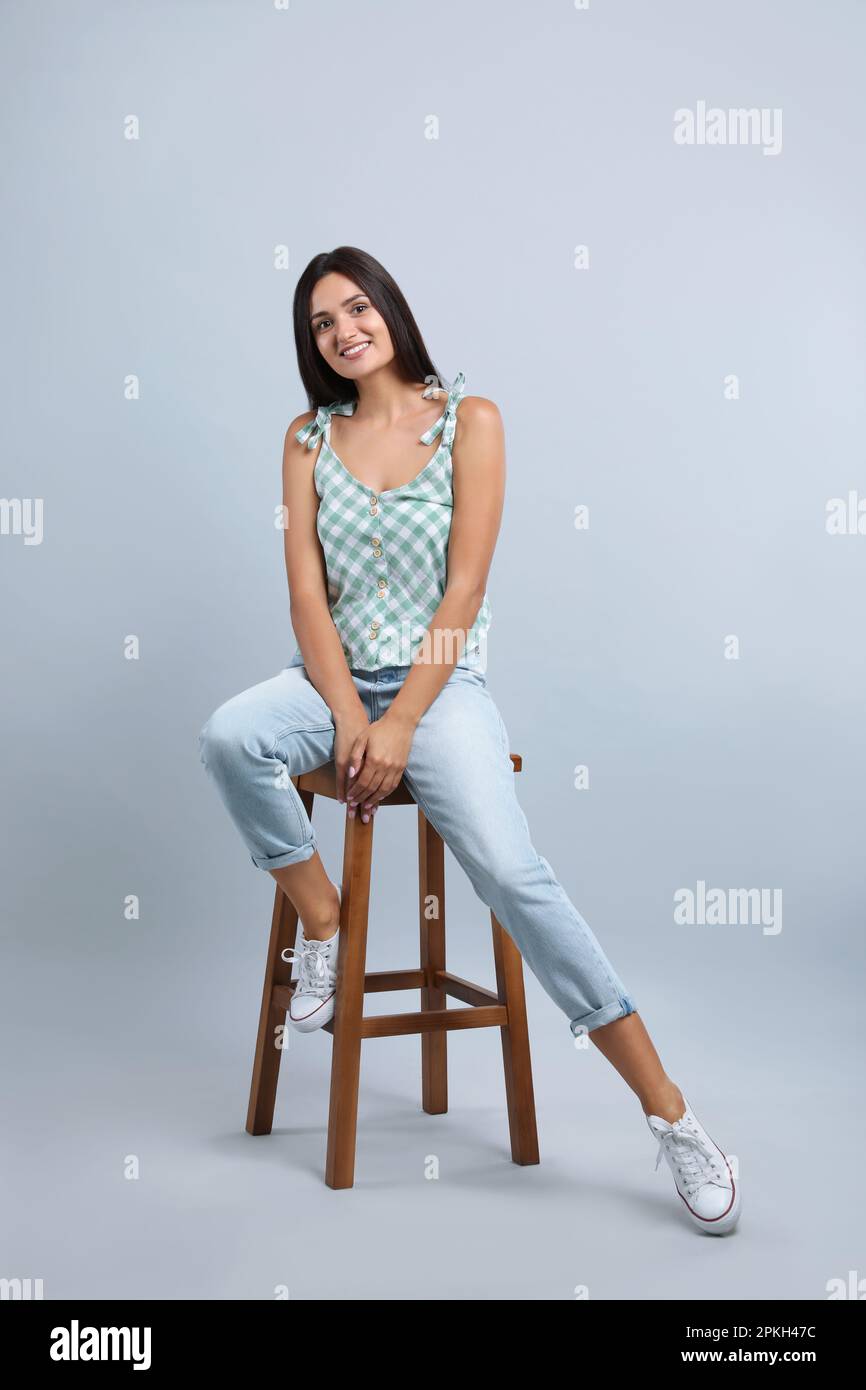 Beautiful young woman sitting on stool against light grey background ...
