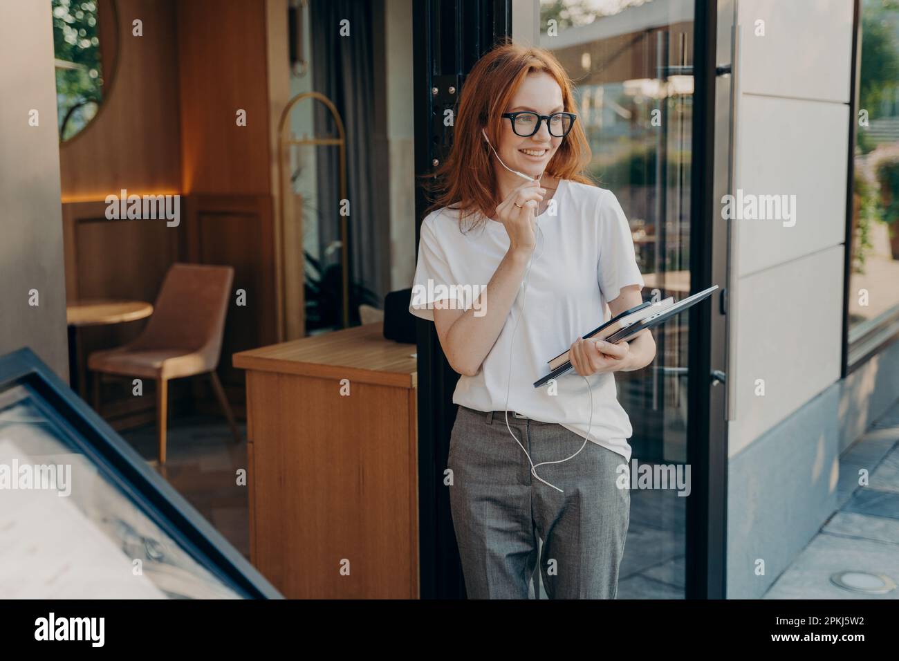 Outdoor shot of glad ginger young woman uses earphones with microphone ...