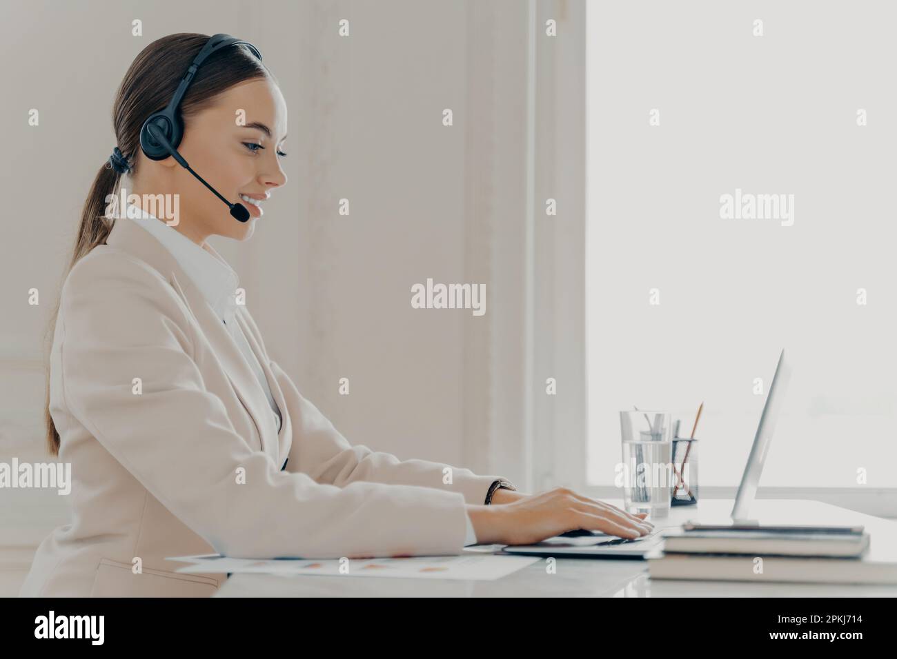 Young friendly female operator in beige formal suit wearing headset and ...