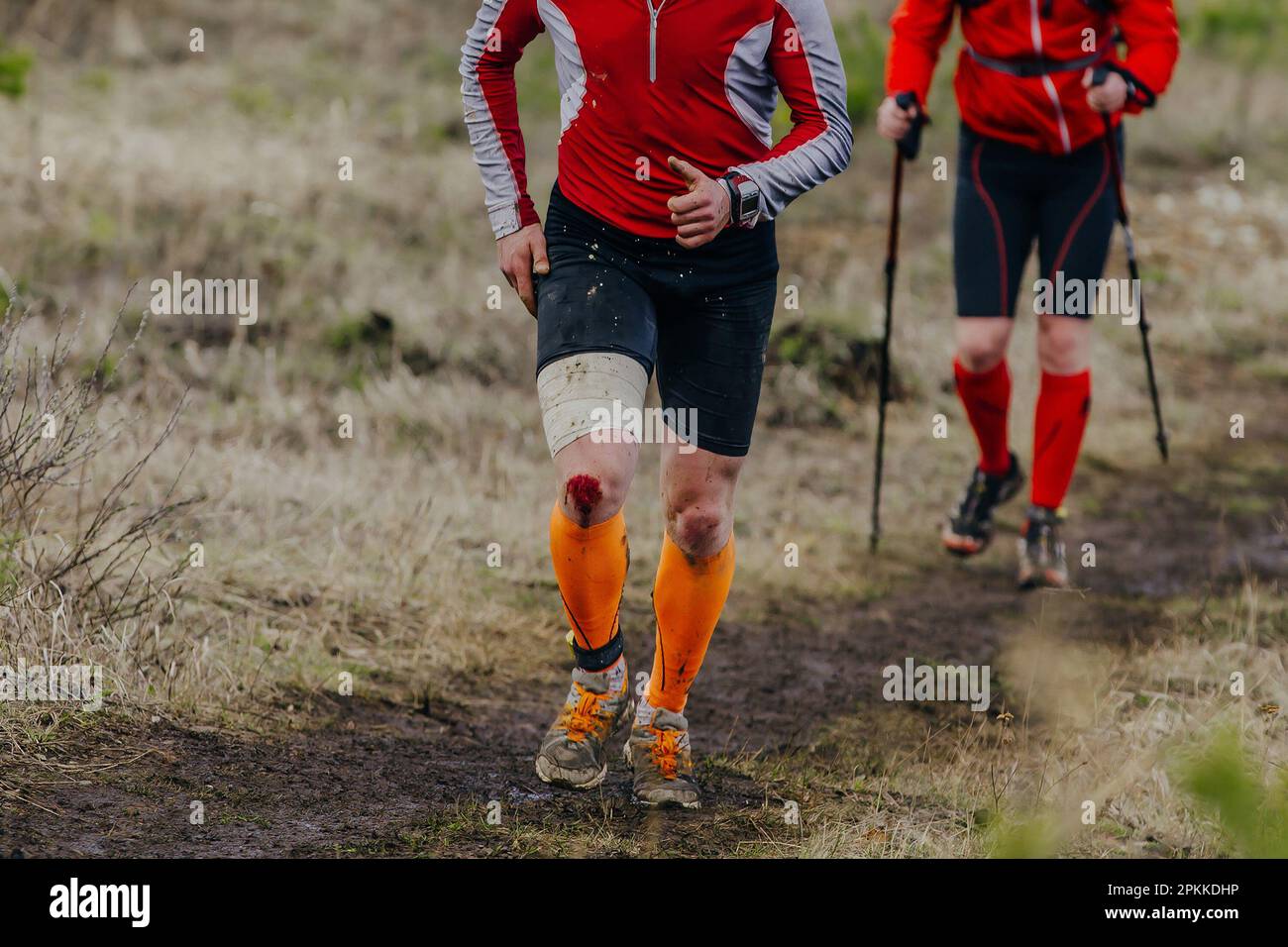 two runners run muddy trail sports race, knee is covered in blood ...
