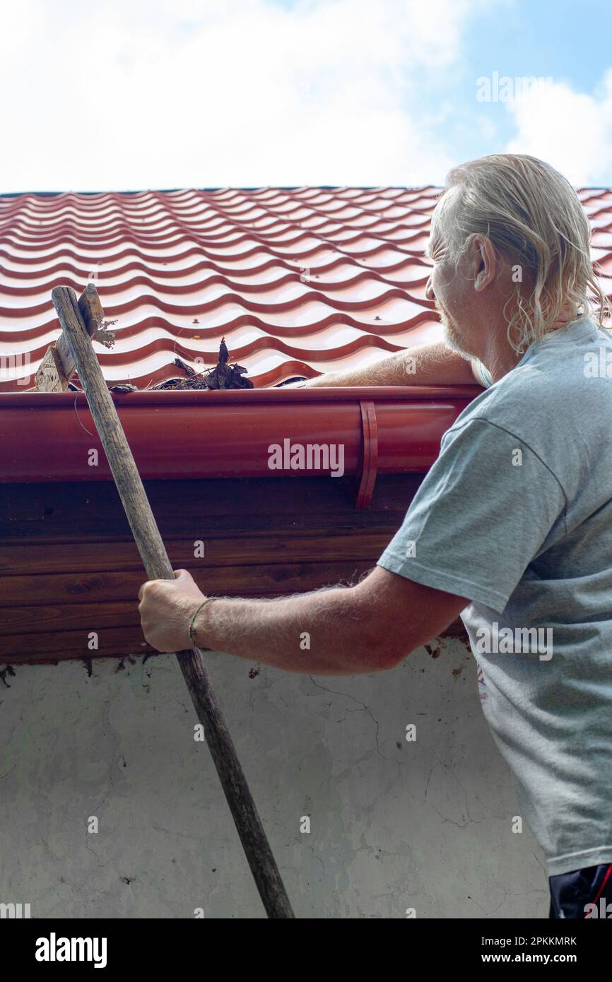 A mature man cleans the gutter of a drainpipe on the roof of his house ...