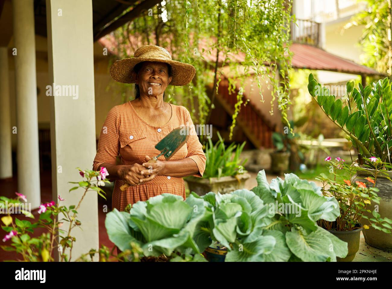 Senior Indian female farmer in straw hat with scoop in garden taking care of potted plants ...